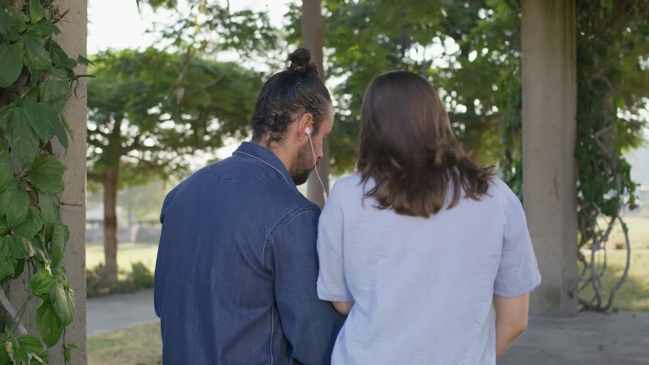 A young couple sits closely on a park bench listening to music sharing ...