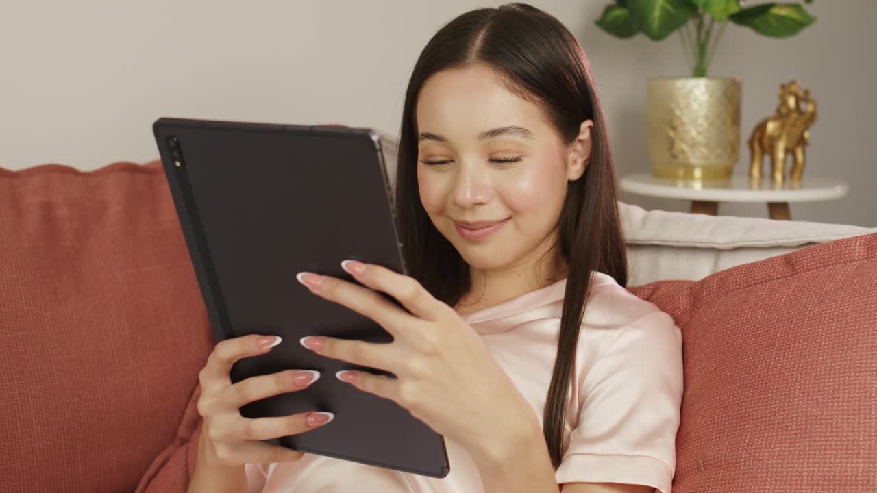A young beautiful woman laying on the couch smiles at the tablet screen ...