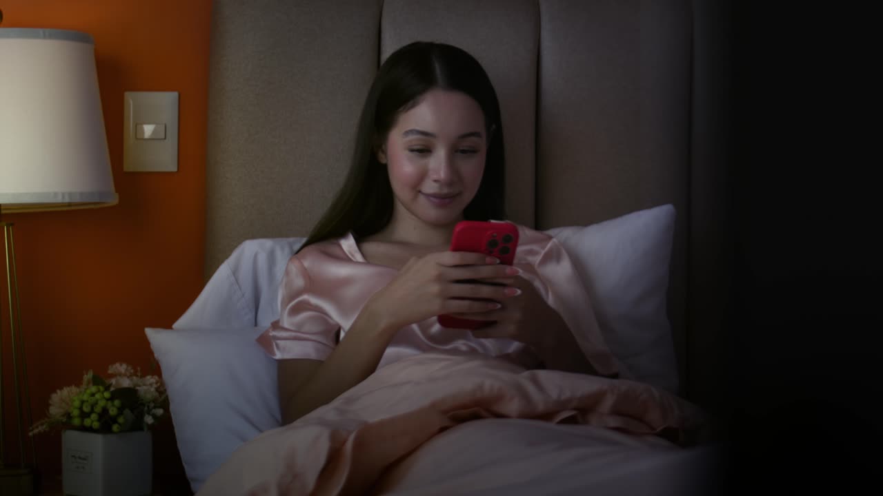 A young smiling woman enjoys social media scrolling on her red phone laying on the comfy bed at ...