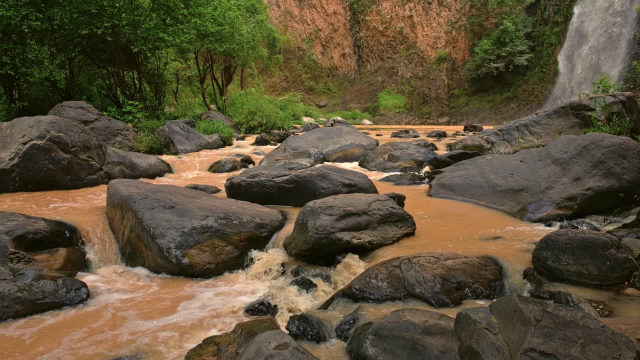 Muddy river flowing through rocky landscape with lush greenery - Free Stock Video