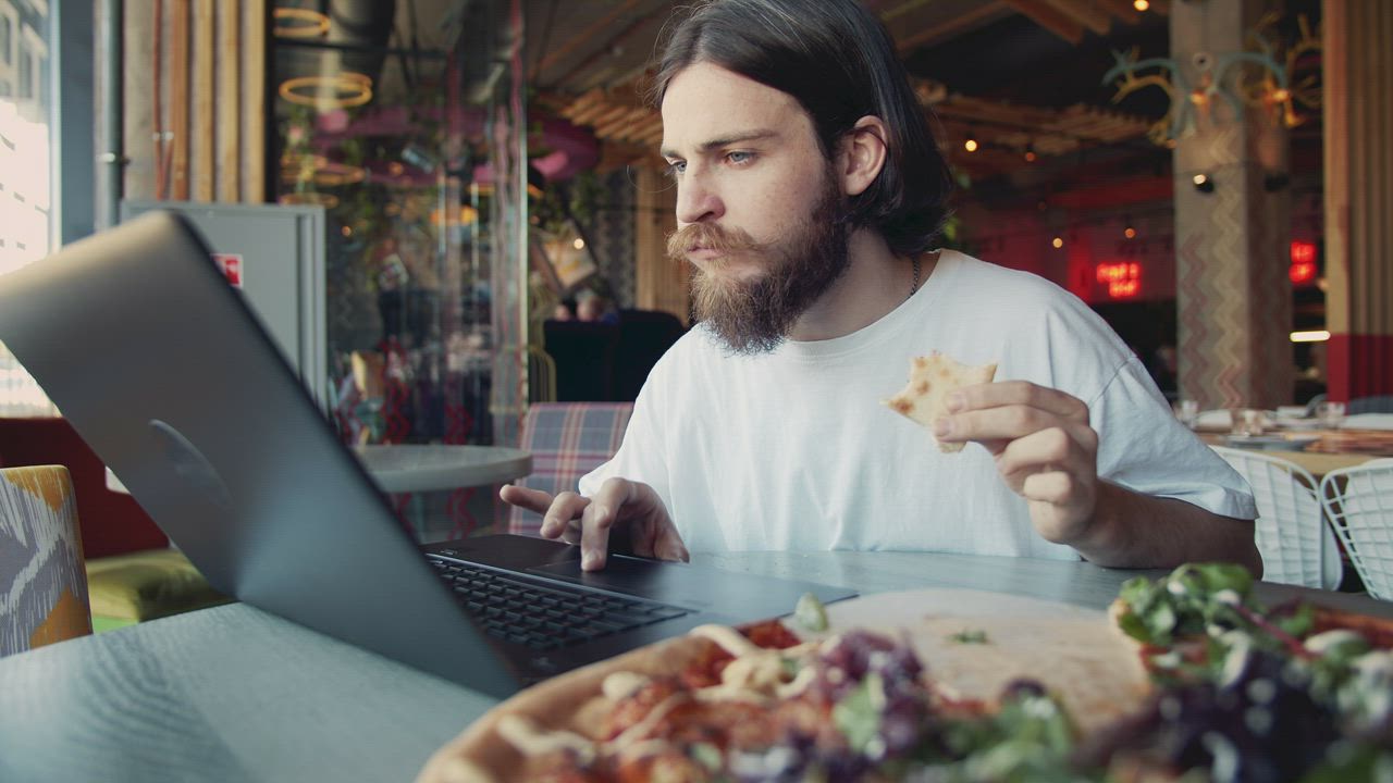 Man eats pizza while woking on laptop at hip cafe - Free Stock Video
