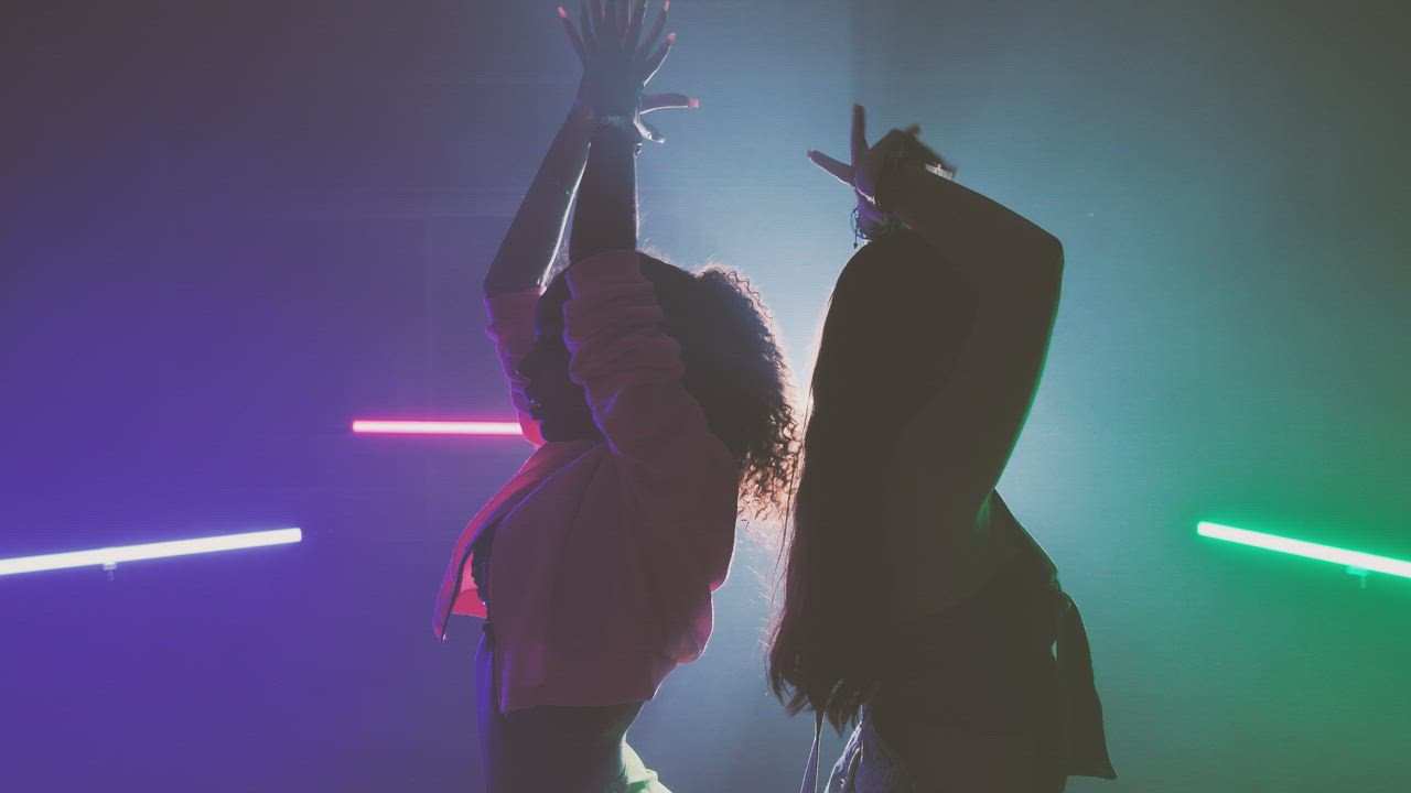 Pair of girls dancing back to back under colored lights - Free Stock Video
