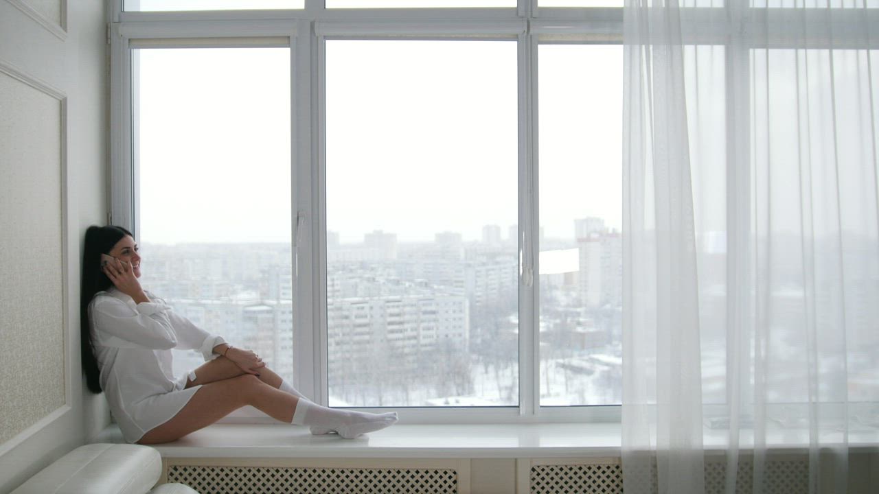Girl sitting at the bottom of a window on a call - Free Stock Video