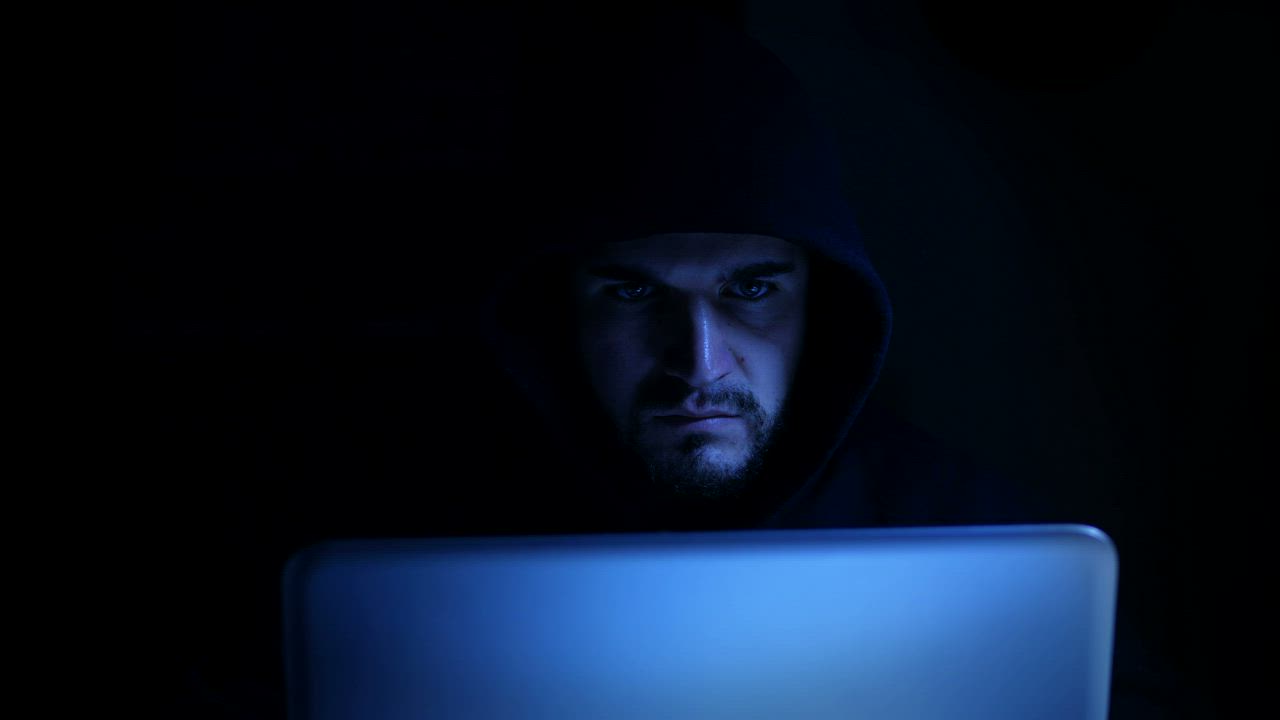 Man wearing a hoodie in a dark room typing aggressively on a laptop ...