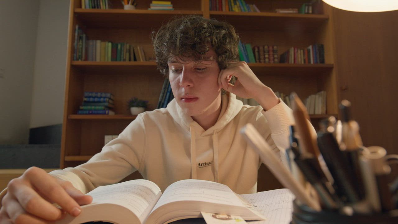 A young student reads information from a book at home with a bookshelf ...