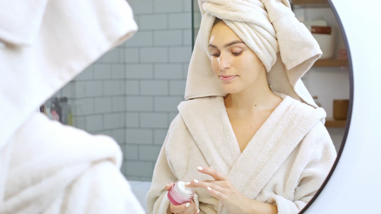 A young woman gently rubs cream on her face in front of the mirror ...