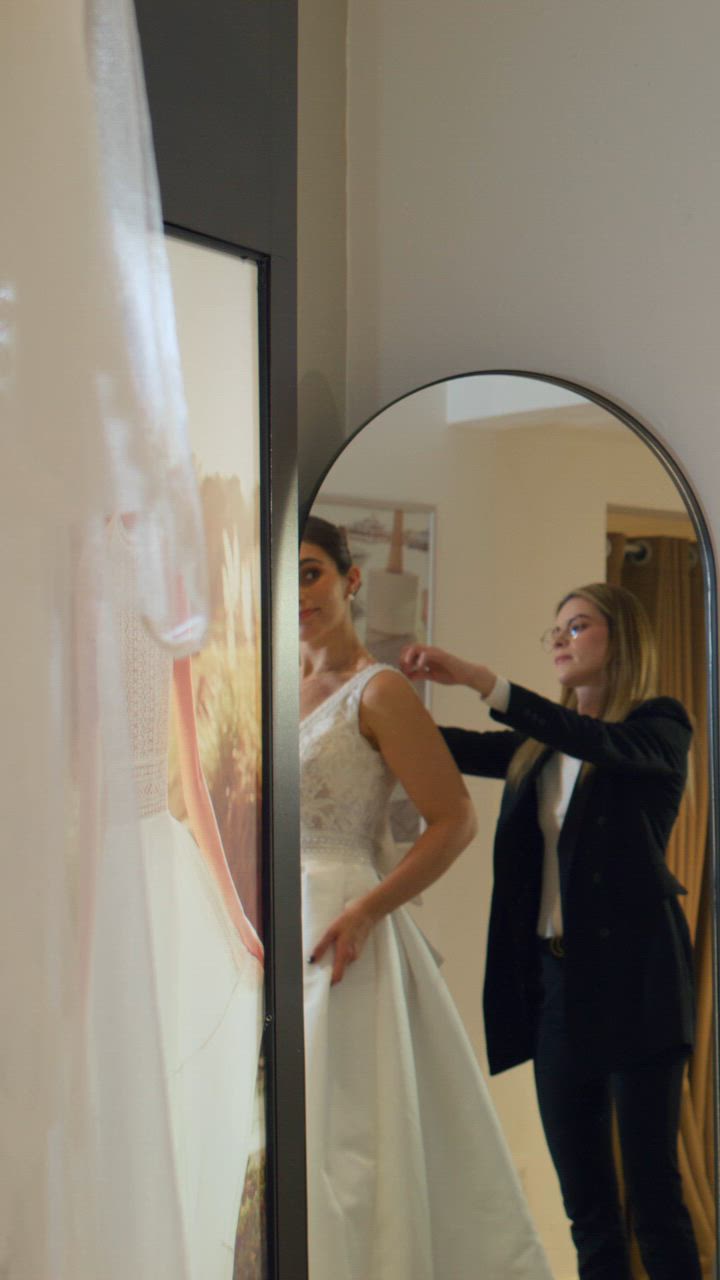 A young beautiful bride stands in front of the mirror as she tries on ...