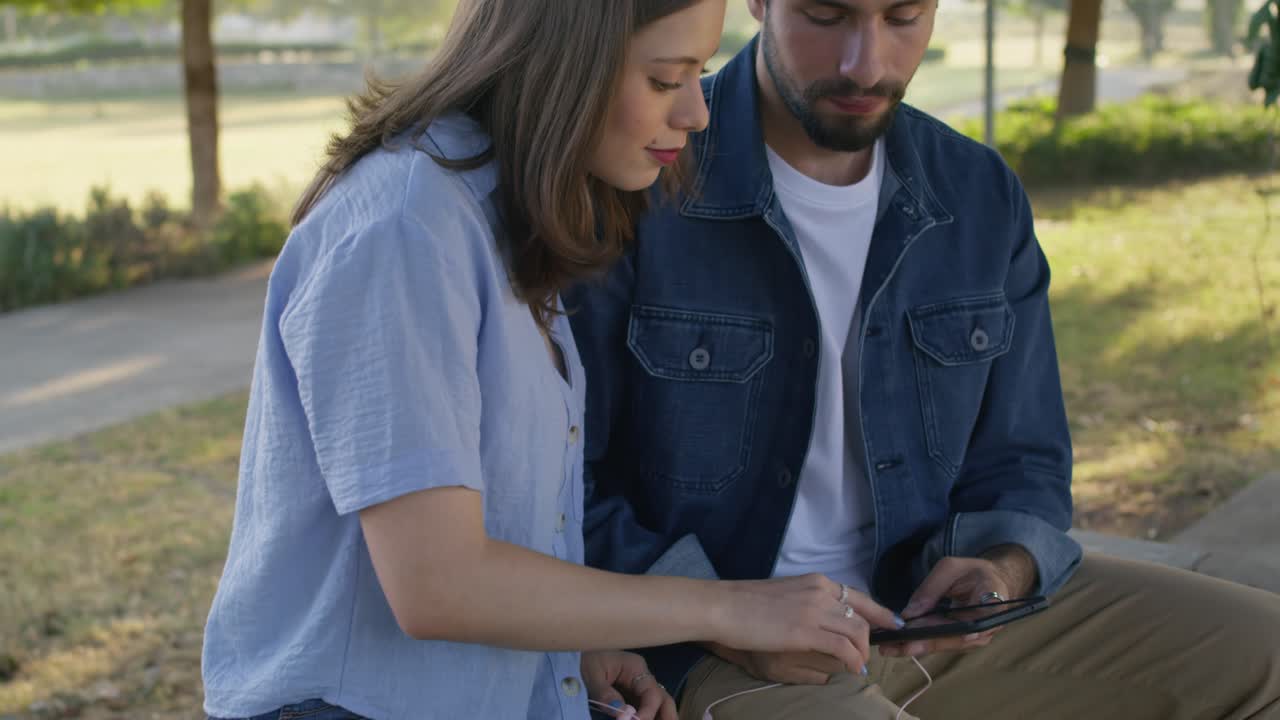 A young couple sitting at the park bench shares a wired headphone and ...